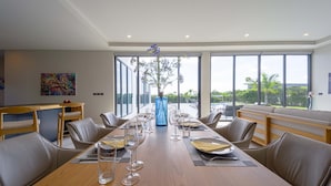 Uma área de jantar moderna com uma longa mesa de madeira posta para refeição, rodeada por cadeiras de couro. Janelas amplas oferecem vista para o exterior.