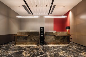 Uma área de recepção de hotel com piso de mármore, balcão de atendimento, cortina vermelha e abajur preto.