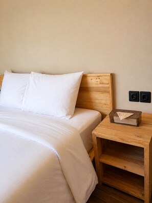 Cama bem arrumada com lençóis e travesseiros brancos, cabeceira de madeira e mesa de cabeceira com um livro e um estojo de lenços.