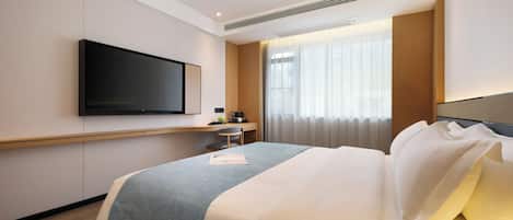 Quarto de hotel com cama, TV de tela plana na parede, mesa pequena com cafeteira e janela com cortinas.