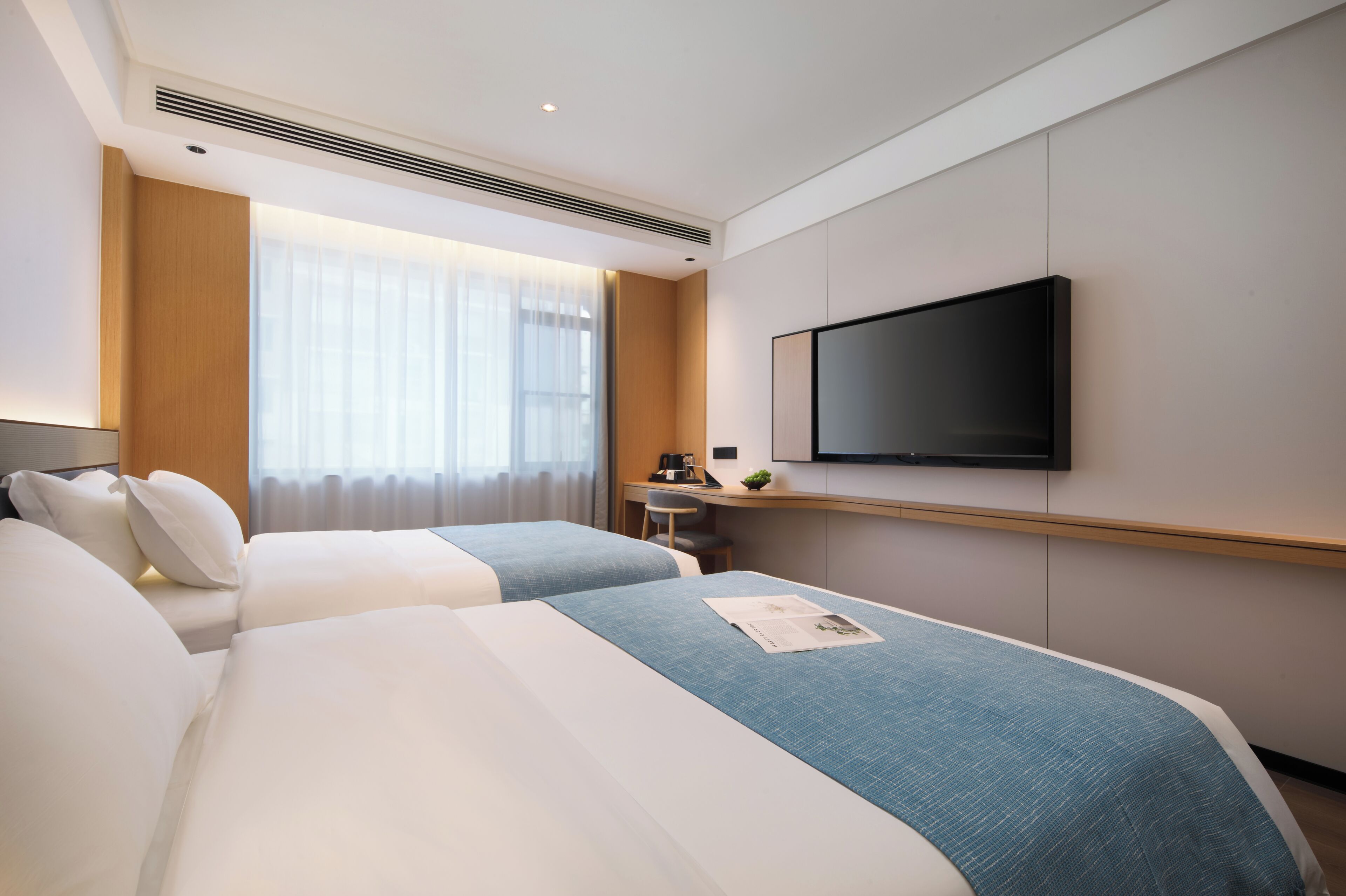 Quarto de hotel com duas camas, televisão de tela plana na parede, escrivaninha com cadeira e janela com cortinas finas.