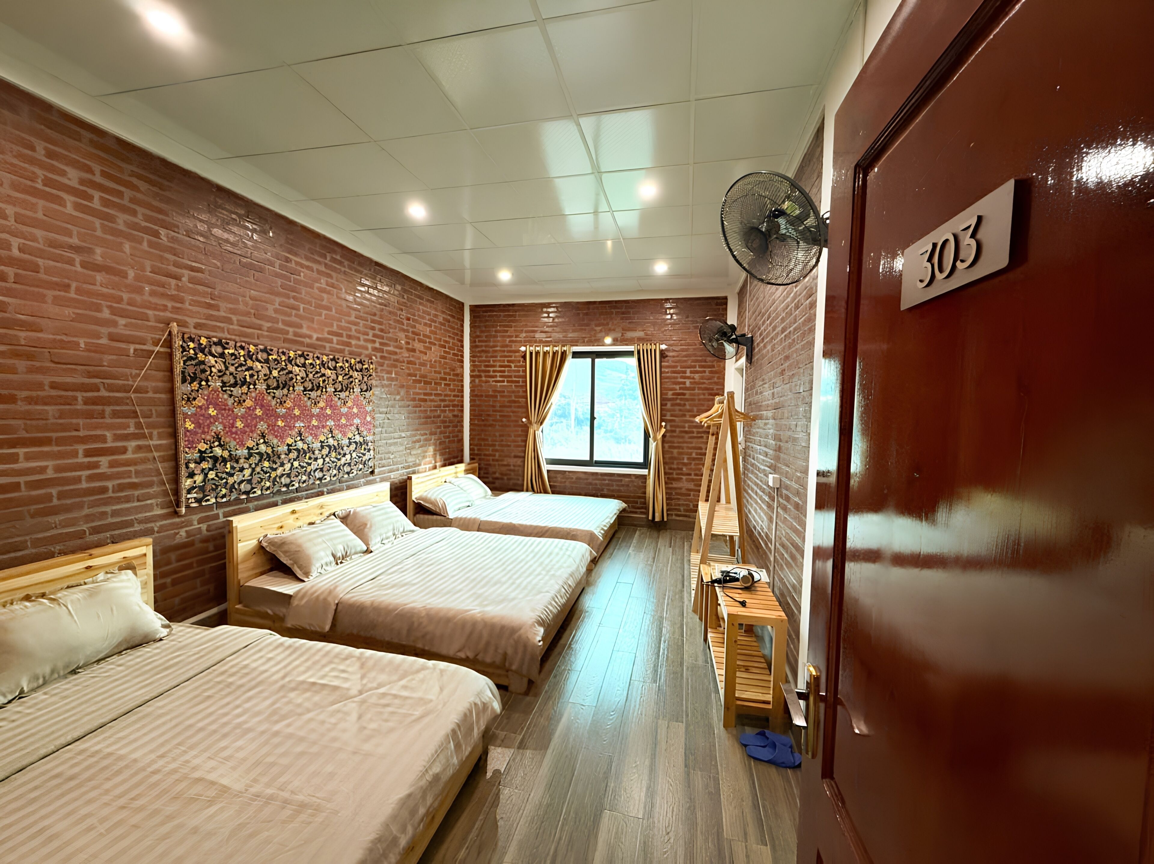 Quarto de hotel com duas camas, uma mesa de madeira, um ventilador, uma janela com cortinas e uma parede de tijolos.