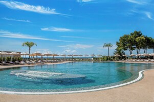 Uma área de piscina com espreguiçadeiras e guarda-sóis, cercada por árvores e um céu claro.