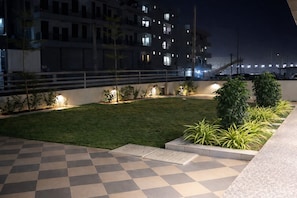 Vista noturna de um pátio com piso xadrez, área gramada e um edifício com janelas iluminadas.