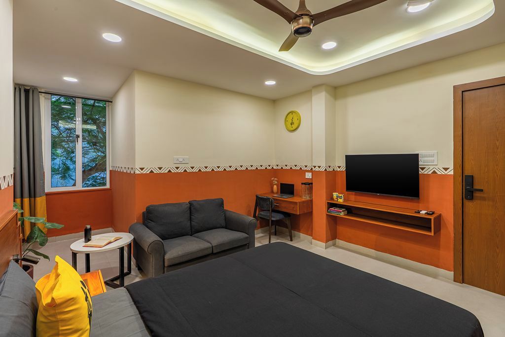 Quarto de hotel com cama, sofá, escrivaninha e computador, televisão e janela com cortinas.