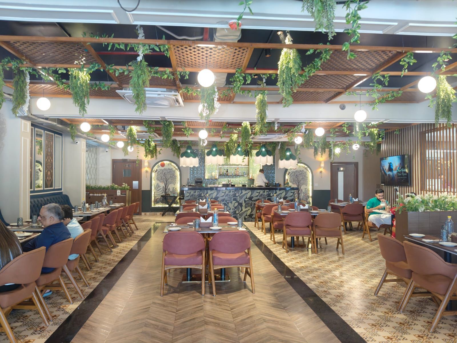 Restaurante com piso em padrão losango, vigas de madeira no teto, plantas penduradas e área de bar com bancada de pedra.