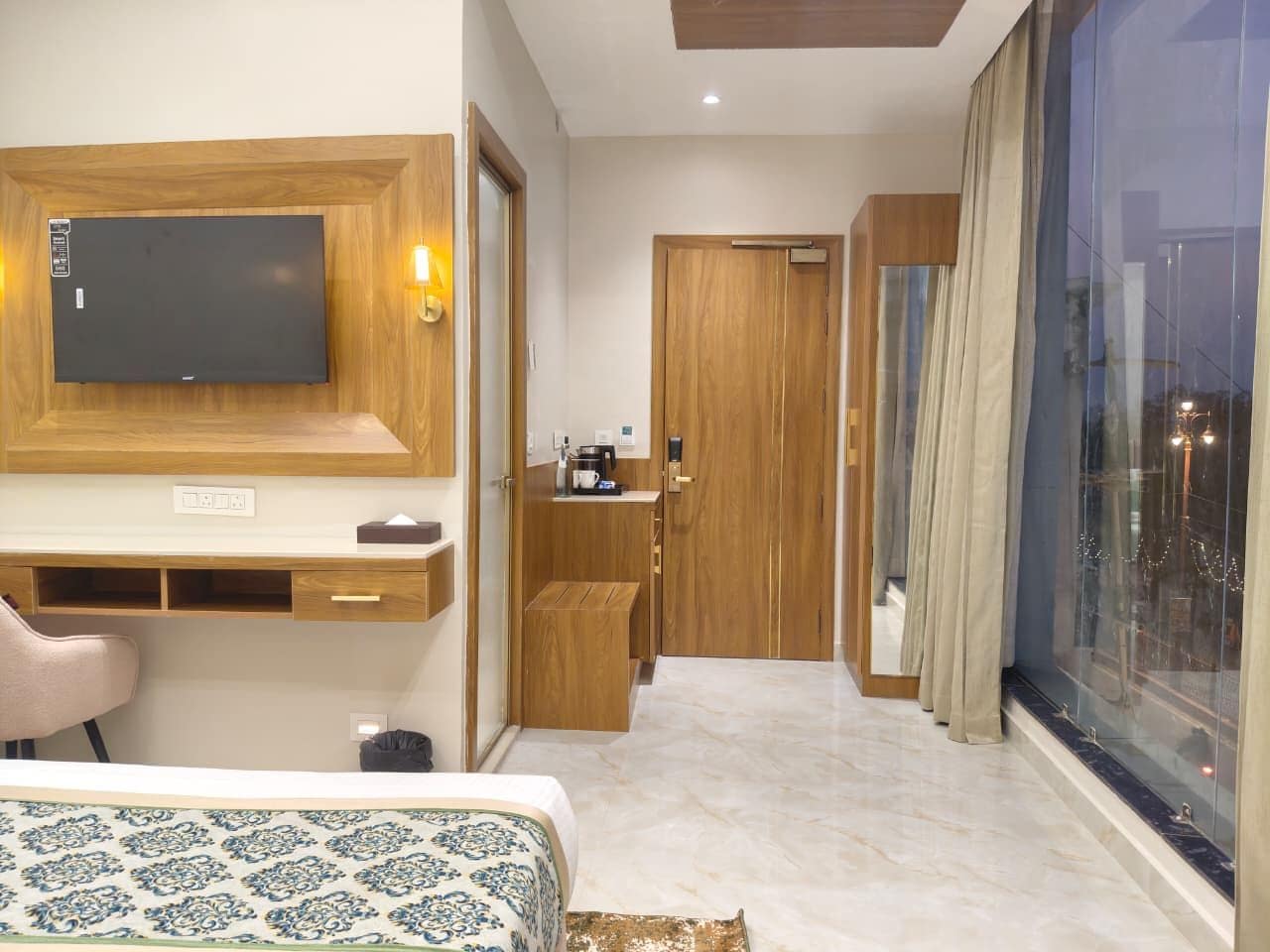 Quarto de hotel com cama, escrivaninha e cadeira, televisão na parede, pequena cozinha e uma janela grande com cortinas.