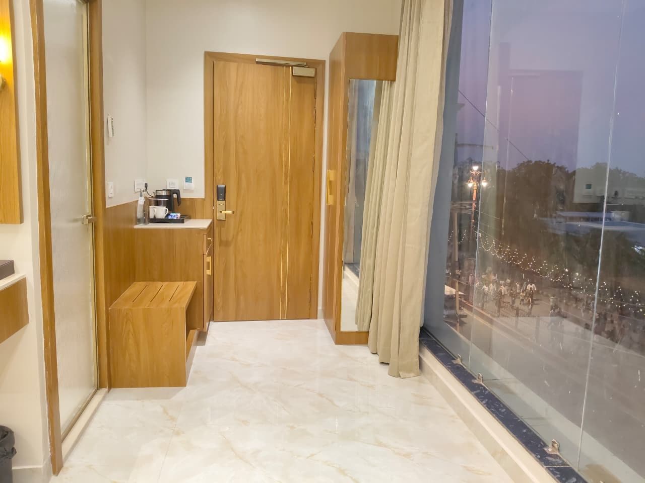 Quarto de hotel com porta de madeira, um banco, uma mesinha com cafeteira e uma janela ampla com vista para a cidade.