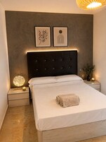 Quarto com cama, mesa de cabeceira com abajur, luminária na parede e duas obras de arte emolduradas.