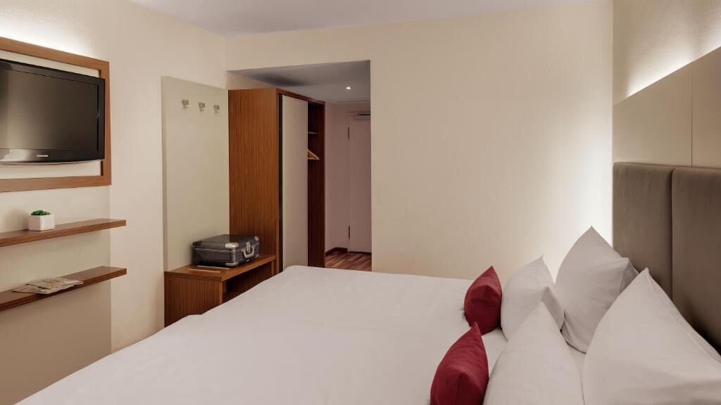 Quarto de hotel com cama, televisão na parede, mesa de cabeceira de madeira e guarda-roupa.