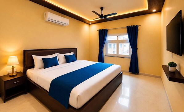 Quarto de hotel com cama, mesa de cabeceira, luminária, ar condicionado, ventilador de teto, janela com cortinas e televisão de tela plana.