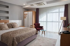 Quarto de hotel moderno com uma cama grande, uma poltrona vermelha, uma mesa pequena e uma escrivaninha com abajur e telefone.