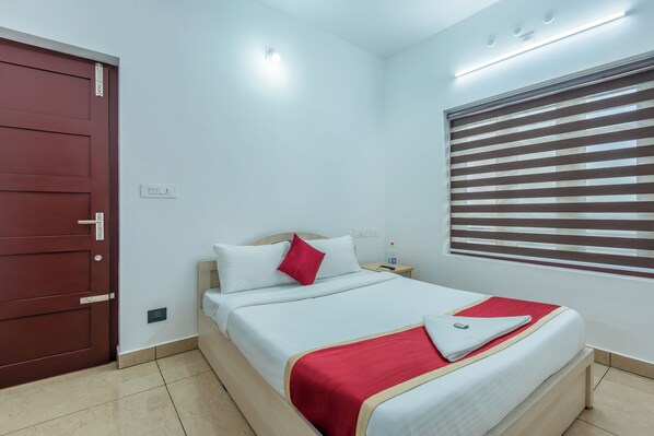 Quarto de hotel com cama, mesa de cabeceira, uma garrafa e janela com persianas.
