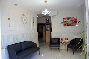 Um hall de hotel com um sofá preto, duas poltronas, uma mesa pequena com vasos e uma grande pintura abstrata, além de um relógio de parede decorativo.