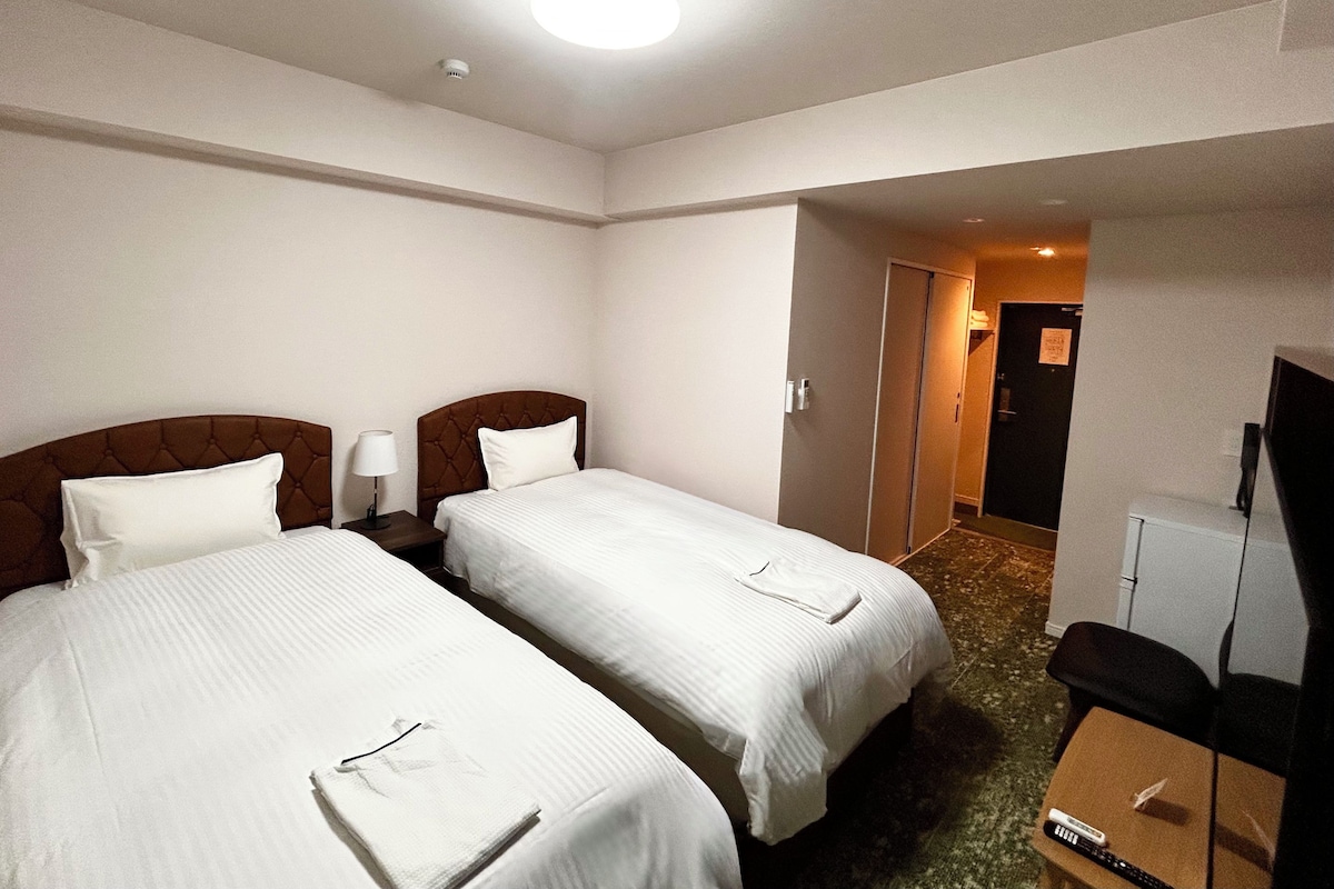 Quarto de hotel com duas camas, uma escrivaninha, uma cadeira, uma televisão e porta que dá para um corredor.