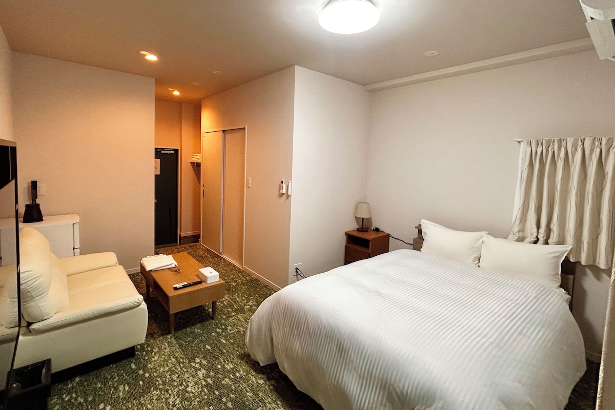 Quarto de hotel com cama, cadeira, escrivaninha, abajur e televisão.