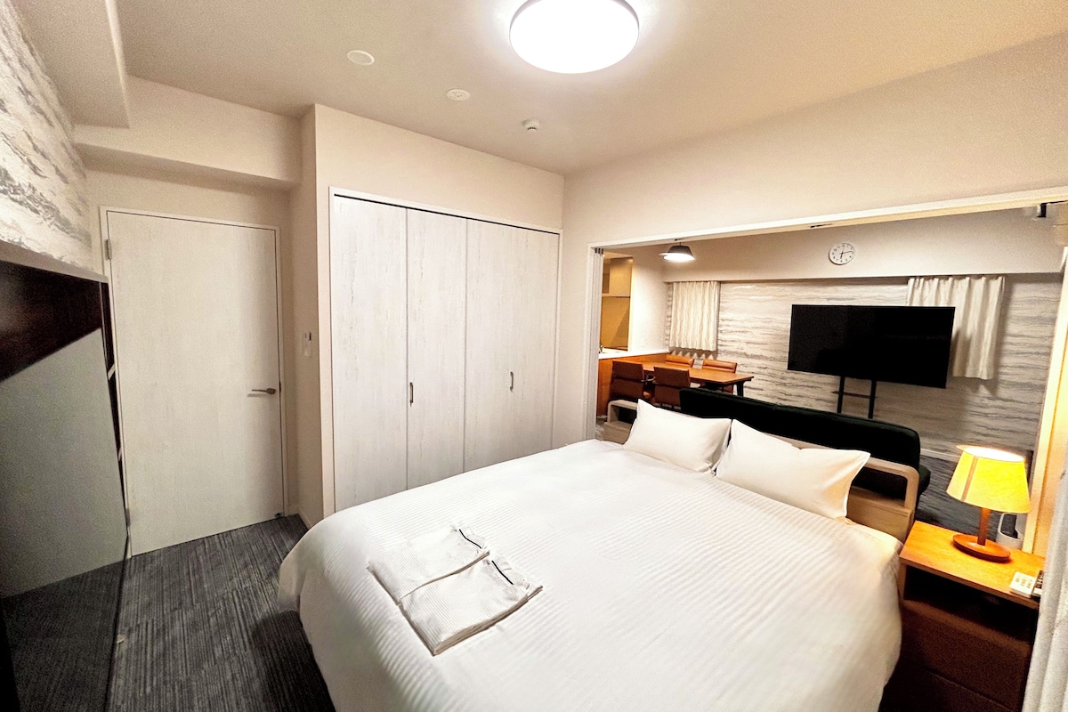 Quarto de hotel com cama, escrivaninha iluminada por uma luminária, televisão na parede e guarda-roupa.