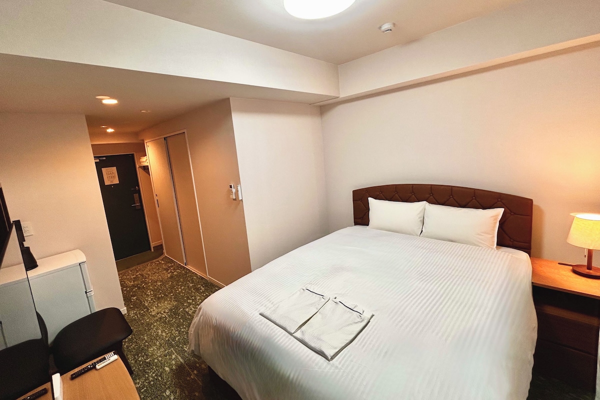 Quarto de hotel com cama, escrivaninha, cadeira, luminária e porta para outro cômodo.