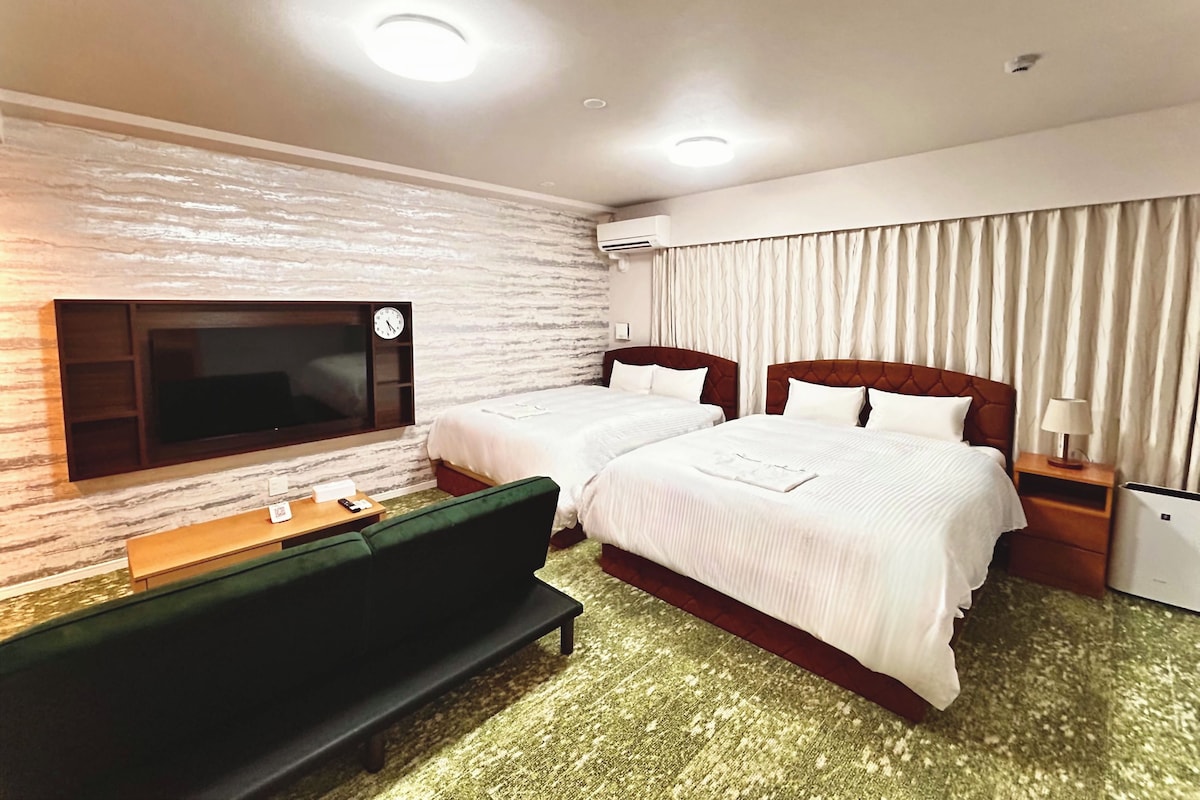 Quarto de hotel com duas camas, televisão na parede, sofá verde, mesa de cabeceira com abajur e piso coberto por carpete.
