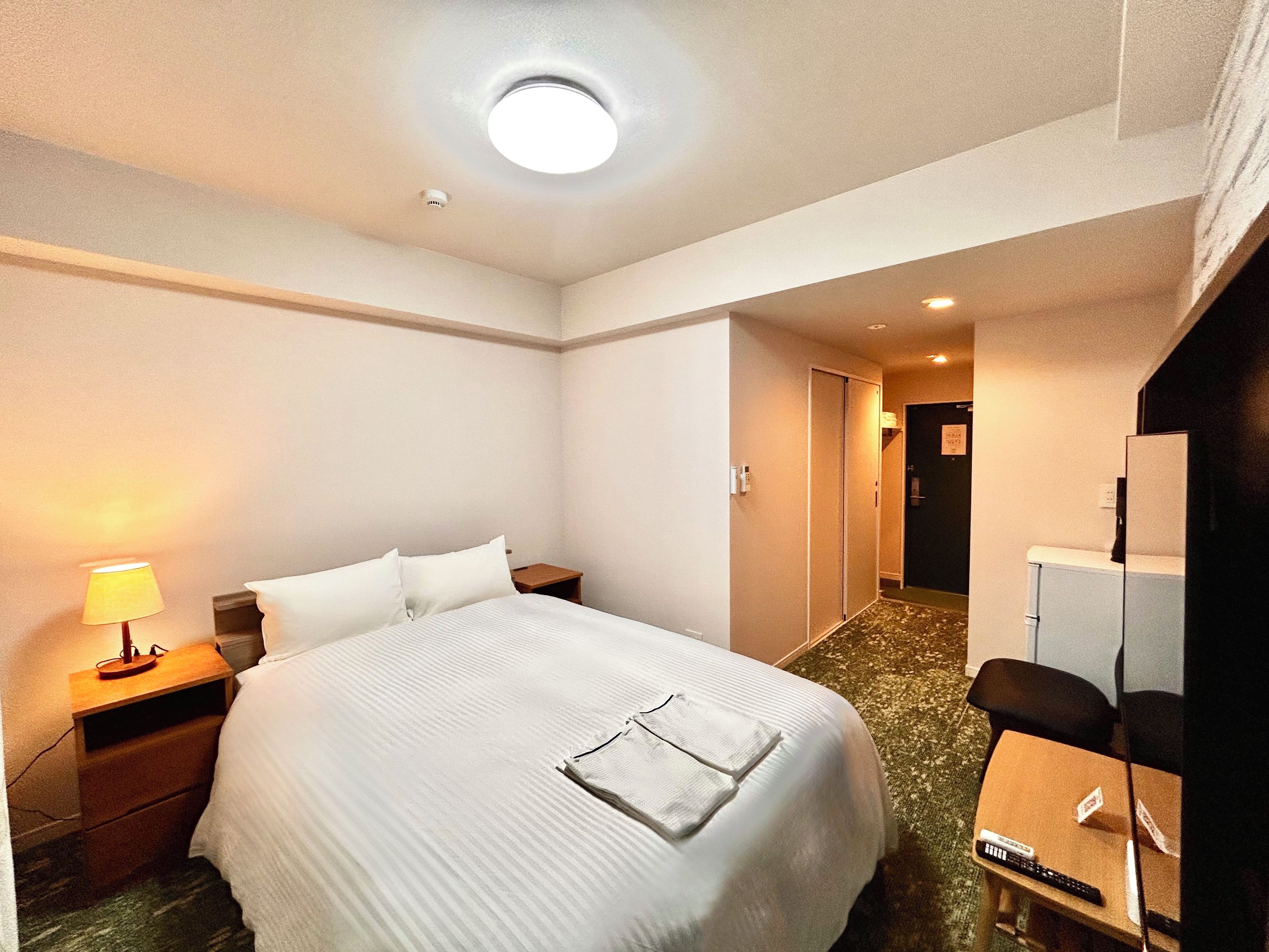 Quarto de hotel com cama, mesa de cabeceira, luminária e escrivaninha com cadeira.