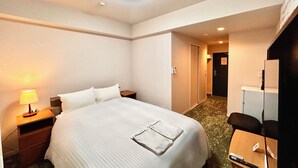 Quarto de hotel com cama, mesa de cabeceira, luminária e escrivaninha com cadeira.
