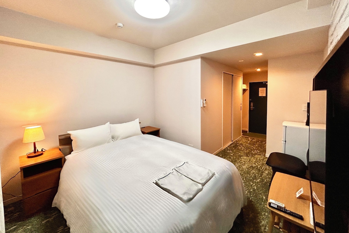 Quarto de hotel com cama, mesa de cabeceira, luminária e escrivaninha com cadeira.