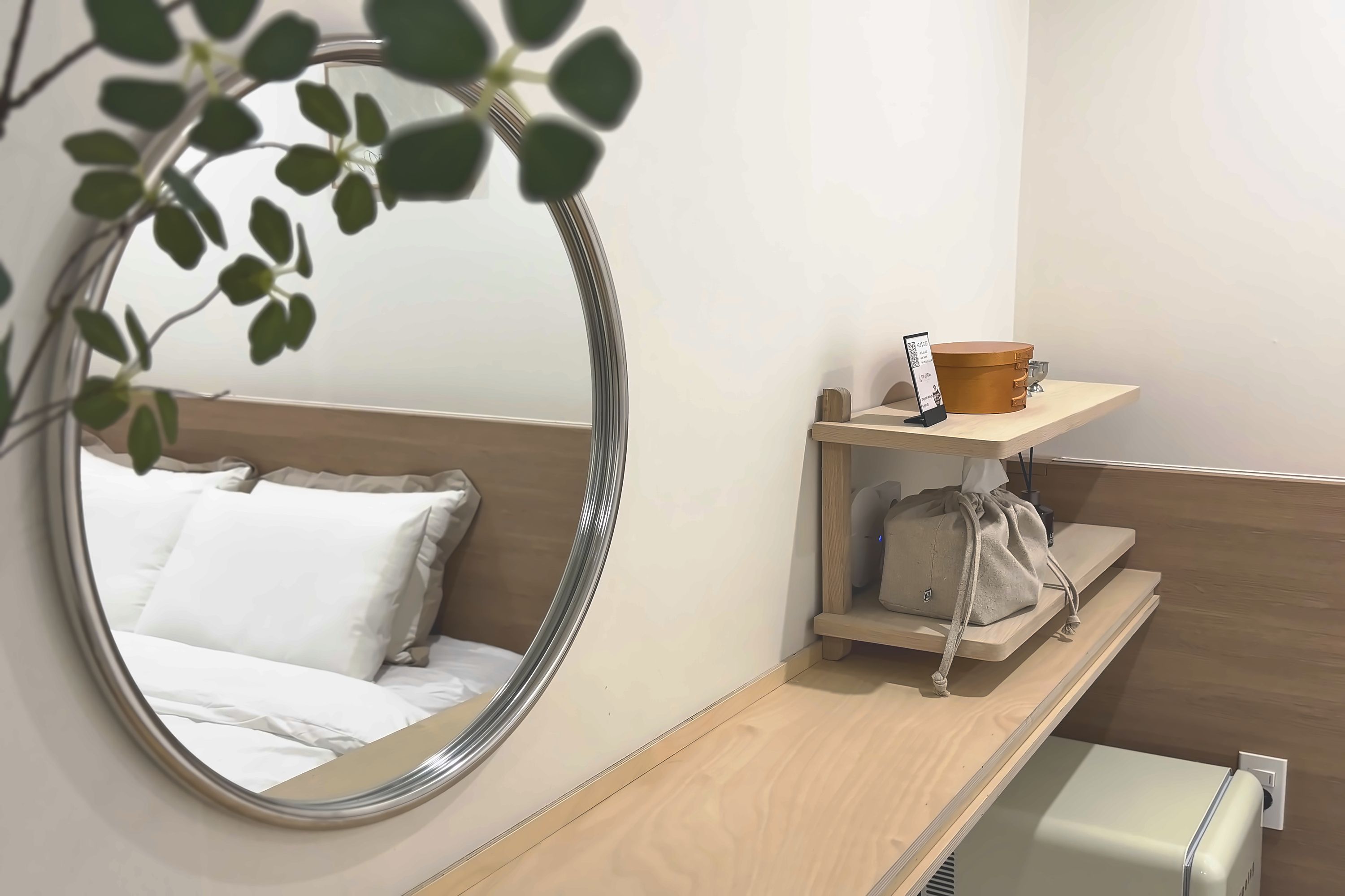 Quarto de hotel com uma cama, um espelho redondo, uma pequena prateleira de madeira com uma xícara, uma bolsa e um pequeno aquecedor.