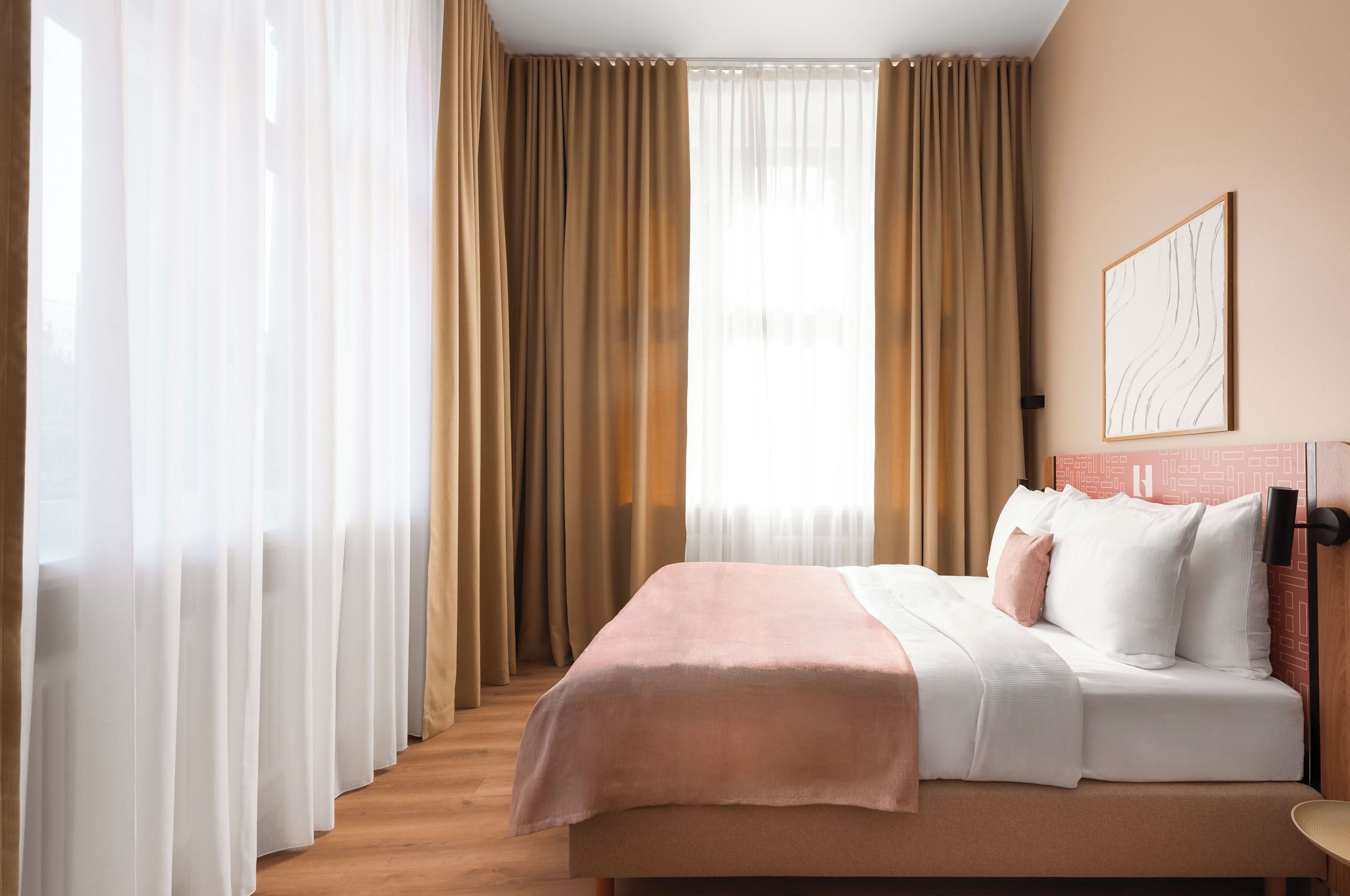Quarto de hotel com cama, cortinas, luminária fixada na parede e quadro emoldurado.