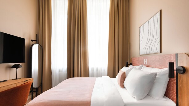 Quarto de hotel com cama, escrivaninha, cadeira, televisão e janela com cortinas.