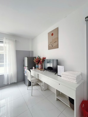 Quarto moderno com escrivaninha branca, televisão de tela plana, geladeira, cadeira, janela com cortinas brancas e uma obra de arte fixada na parede.