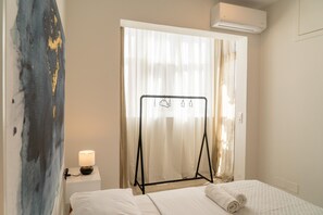 Quarto de hotel com cama, ar condicionado fixado na parede, mesa de cabeceira com luminária e um cabideiro.