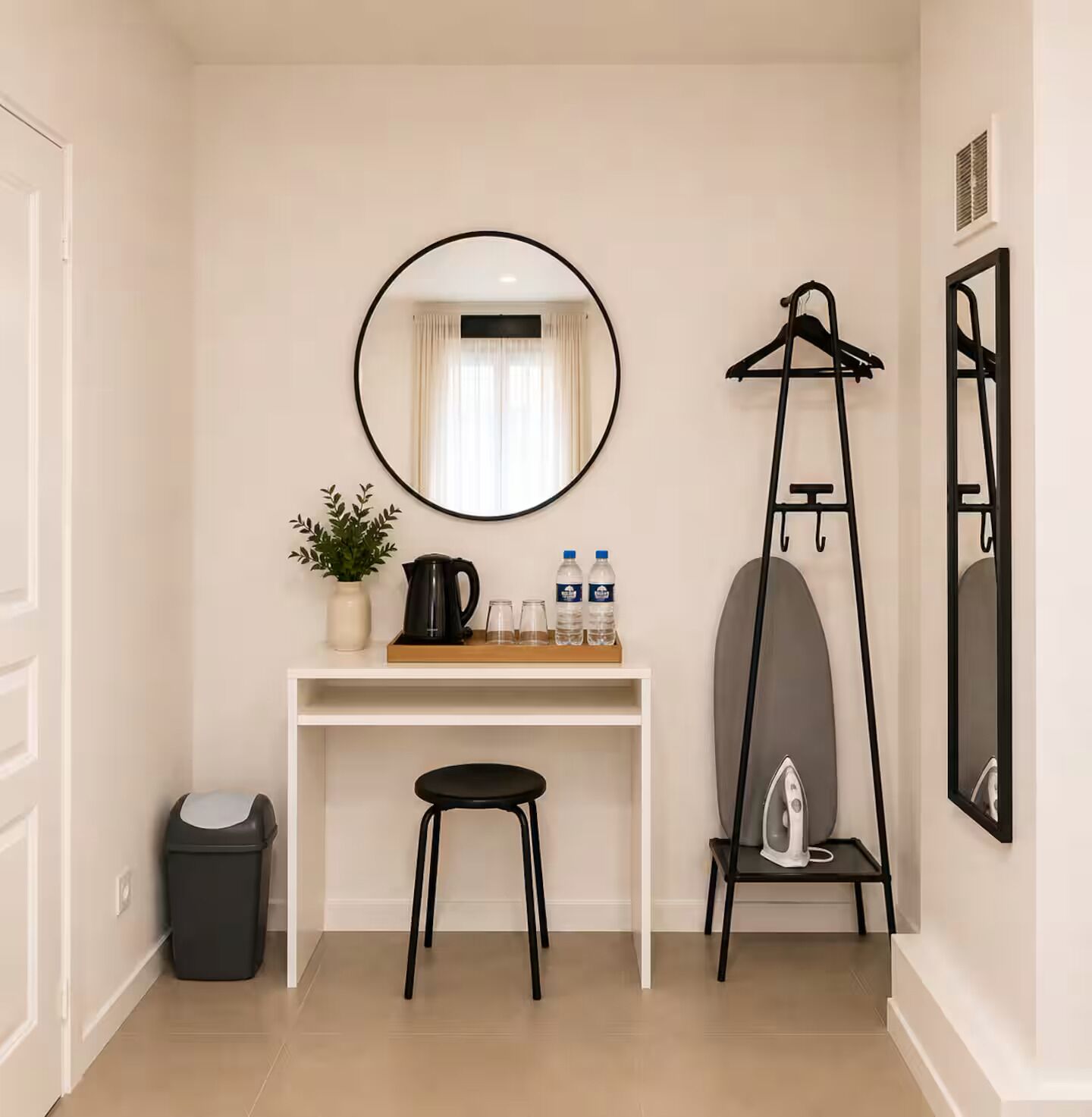 Quarto compacto com escrivaninha branca, espelho redondo, cabide preto, um banquinho pequeno, lixeira e um vaso de planta.