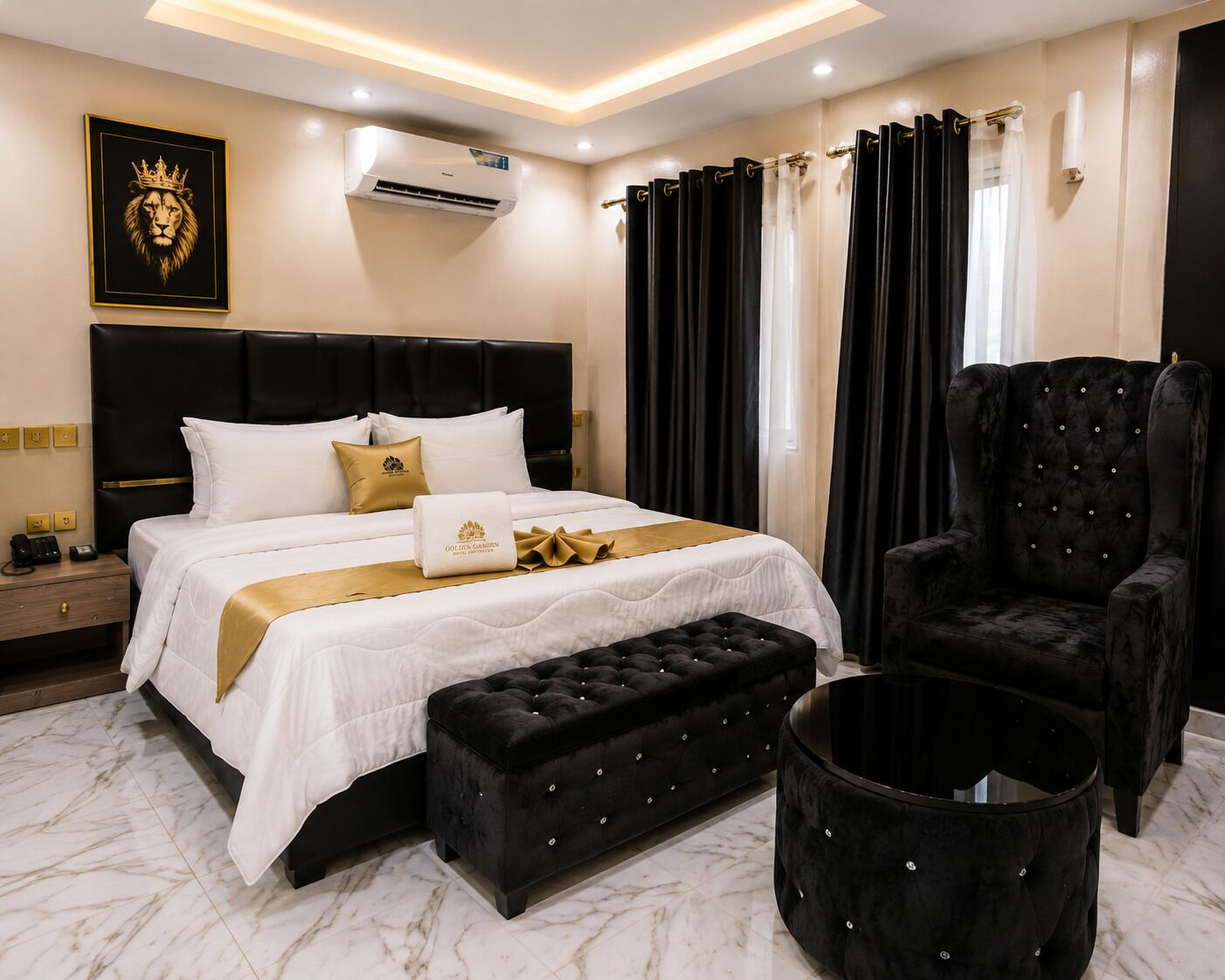 Quarto de hotel com uma cama grande, uma poltrona estofada preta, um banquinho redondo preto, almofada dourada decorativa, um quadro na parede e um ar condicionado.