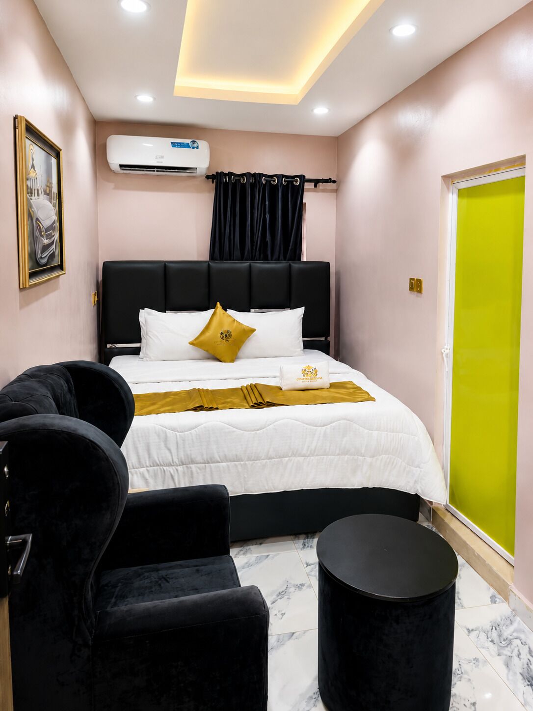 Quarto de hotel com cama, cadeiras estofadas pretas, uma mesa redonda preta, ar condicionado fixado na parede e uma porta verde vibrante.