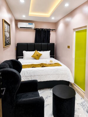 Quarto de hotel com cama, cadeiras estofadas pretas, uma mesa redonda preta, ar condicionado fixado na parede e uma porta verde vibrante.