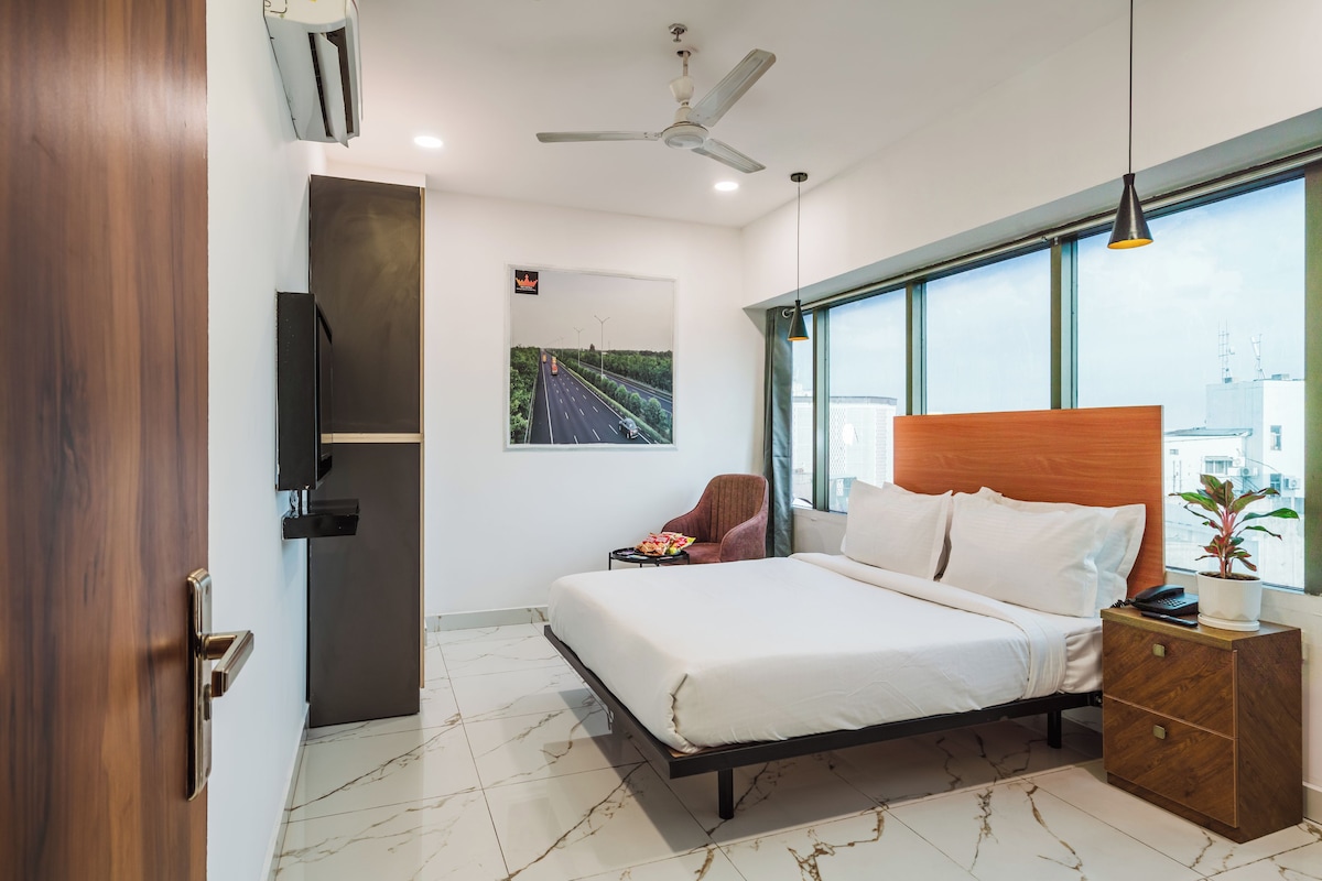 Quarto de hotel com cama, cadeira, mesa de cabeceira, planta em vaso, janela com vista e ventilador de teto.