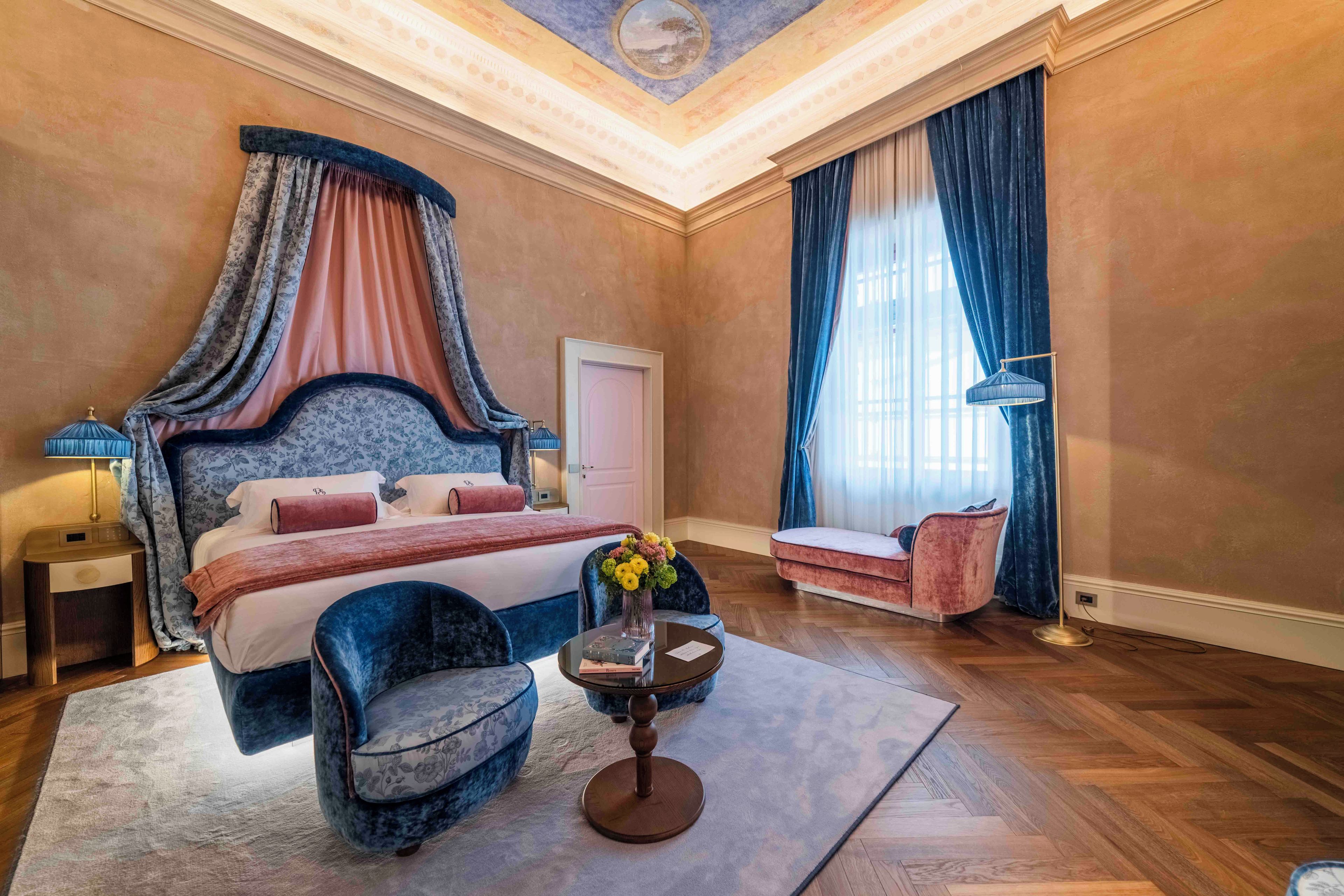 Quarto de hotel com cama de dossel, área de estar com uma cadeira e uma mesa, uma mesinha de cabeceira pequena e uma grande janela com cortinas.