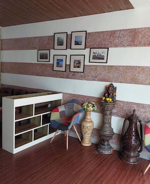 Quarto com piso de madeira, estante branca, cadeira colorida, vaso decorativo e parede com quadros.