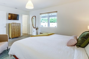 Quarto com cama grande, televisão na parede, janela vista para árvores e porta que dá para outro cômodo.