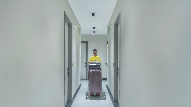 Uma pessoa vestindo uma camisa amarela empurrando um carrinho com garrafas de água por um corredor com portas de ambos os lados.