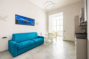 Uma sala de estar moderna com um sofá azul, uma área de jantar com uma mesa e cadeiras brancas, e uma cozinha com forno embutido.