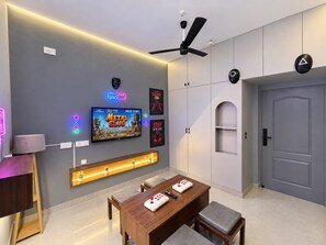 Uma sala de estar moderna com televisão de tela plana, pôsteres de jogos, ventilador de teto e um espaço de jantar com mesa e cadeiras de madeira.