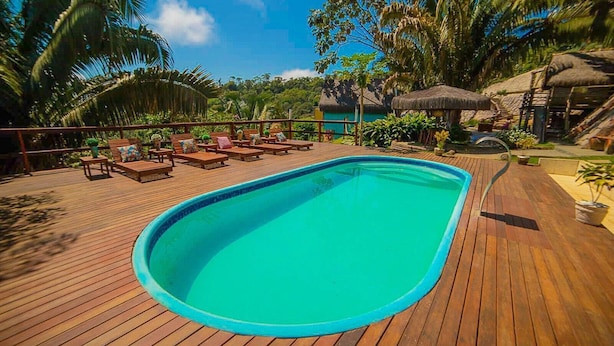 Um deck de madeira com piscina, espreguiçadeiras e estruturas com telhado de palha.