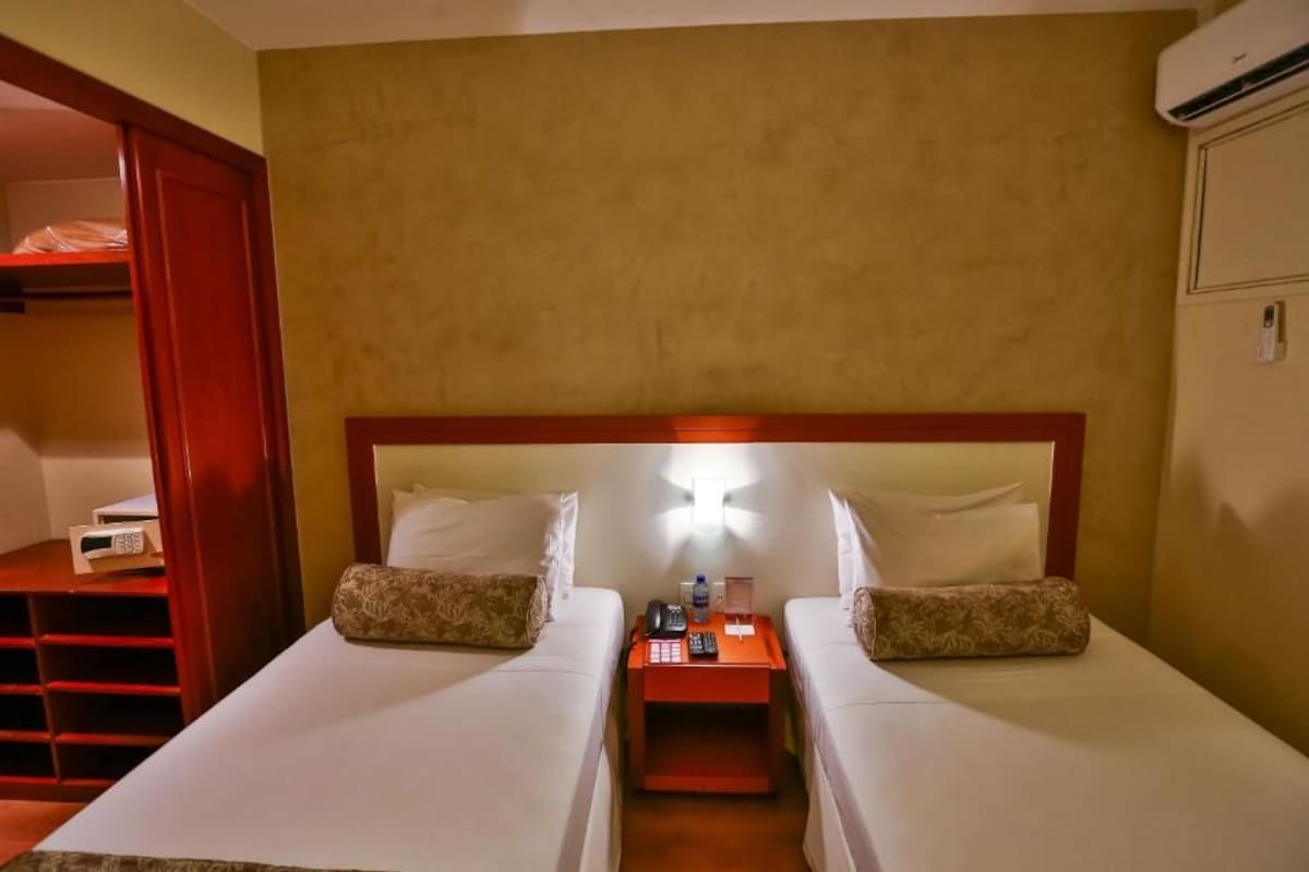 Quarto de hotel com duas camas de solteiro, mesa de cabeceira com abajur e um móvel de armazenamento vermelho.