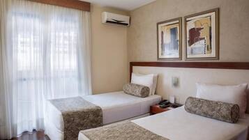 Quarto de hotel com duas camas, uma janela com cortinas leves, um ar-condicionado e duas obras de arte emolduradas na parede.