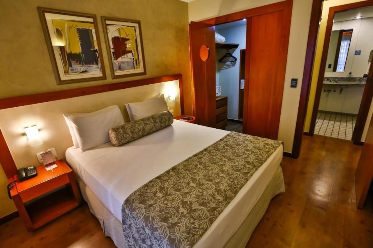 Quarto de hotel com cama, mesa de cabeceira e espelho. Há uma porta que dá para um banheiro e um corredor.