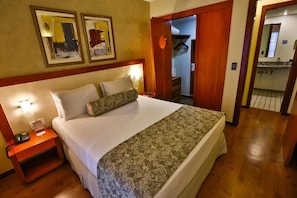 Quarto de hotel com cama, mesa de cabeceira e espelho. Há uma porta que dá para um banheiro e um corredor.