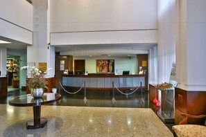 Uma área de recepção de hotel com balcão, sala de espera com mesa e cadeiras, e um vaso decorativo com flores.