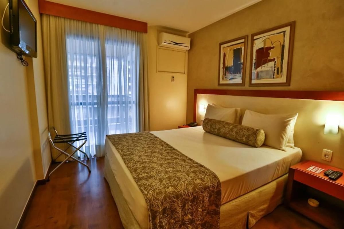 Quarto de hotel com uma cama, mesa de cabeceira, televisão, aparelho de ar condicionado e janela com cortinas.