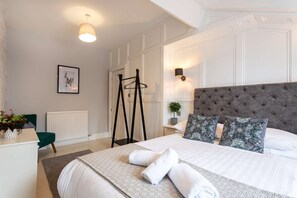 Quarto de hotel com cama, radiador, planta emoldurada, cabideiro e luminária na parede.